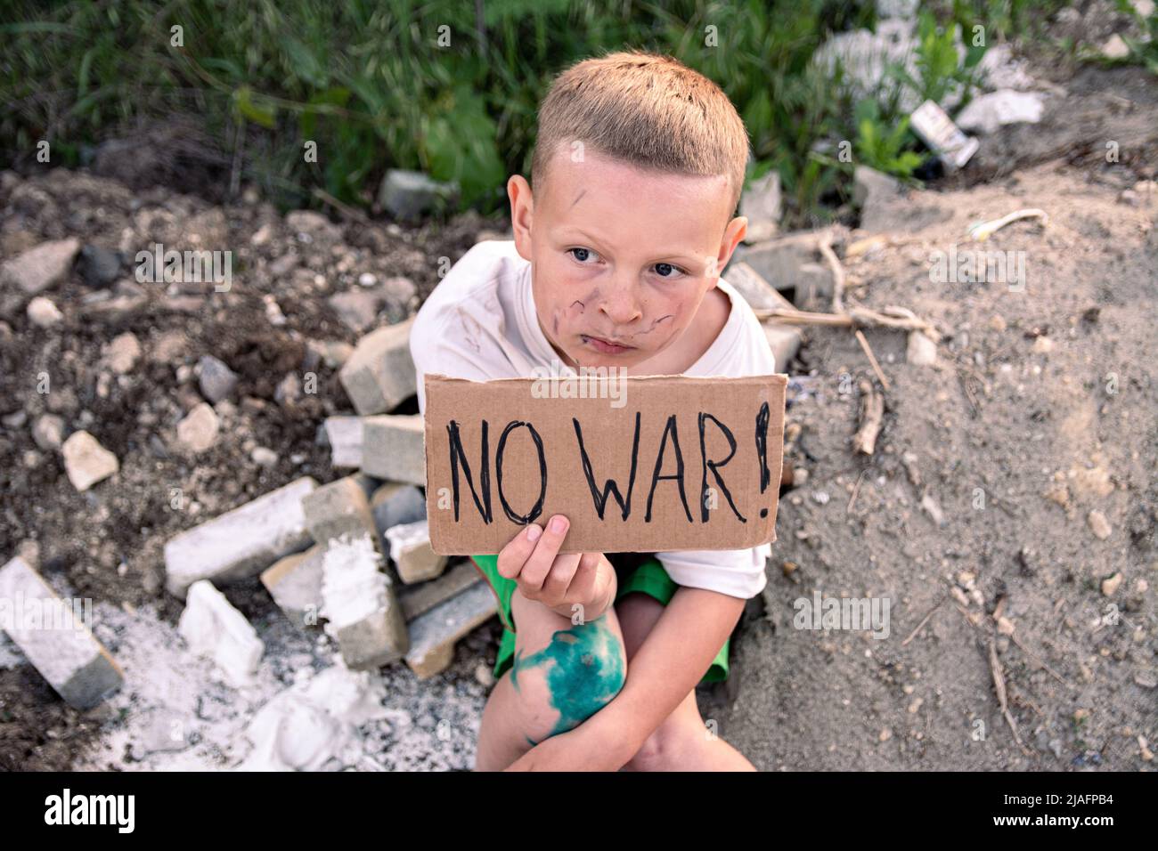 A boy with a grimy face and sad eyes holds a cardboard poster with the ...