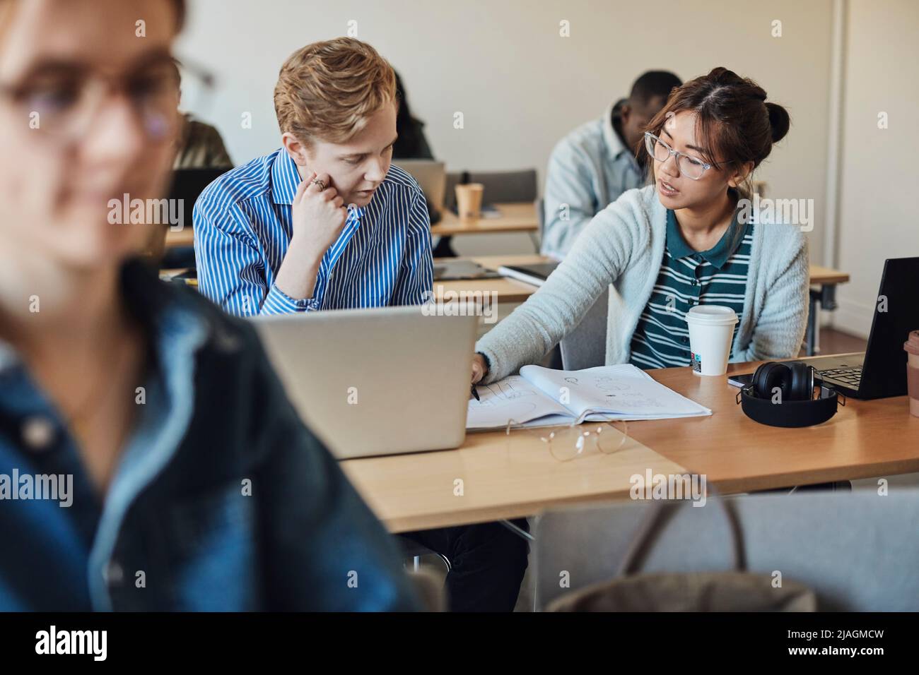 Female student helping male friend while sitting at desk in classroom ...