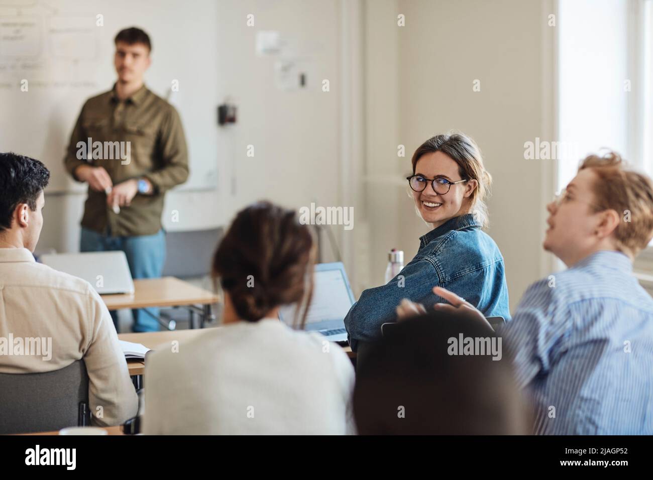Smiling female student looking at friend during lecture in classroom ...