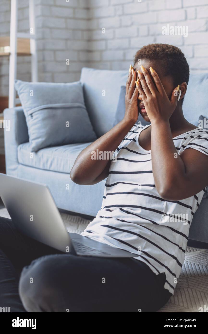 Stressed afro woman grabbing head and using laptop, while sitting on ...