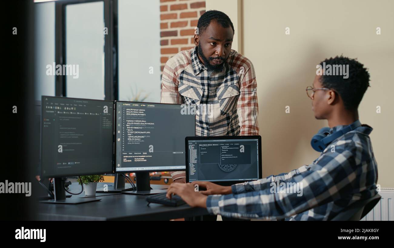 Software developer sitting at desk with multiple screens and laptop running code talking with ...