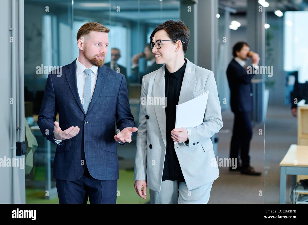 Two young colleagues talking to each other while walking along modern ...