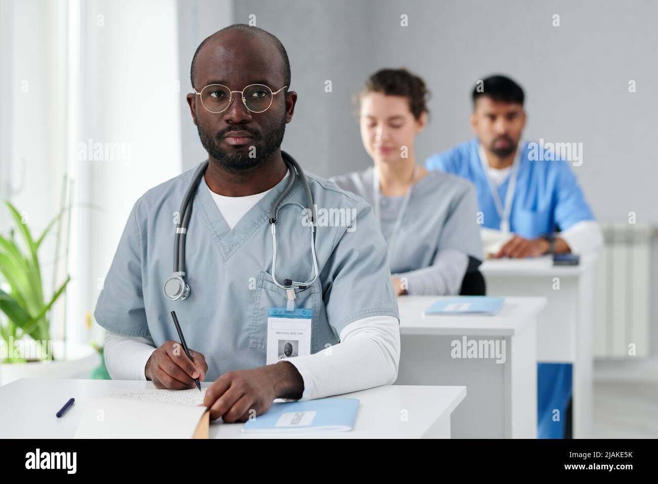 Young African doctor in eyeglasses making notes while sitting at desk ...
