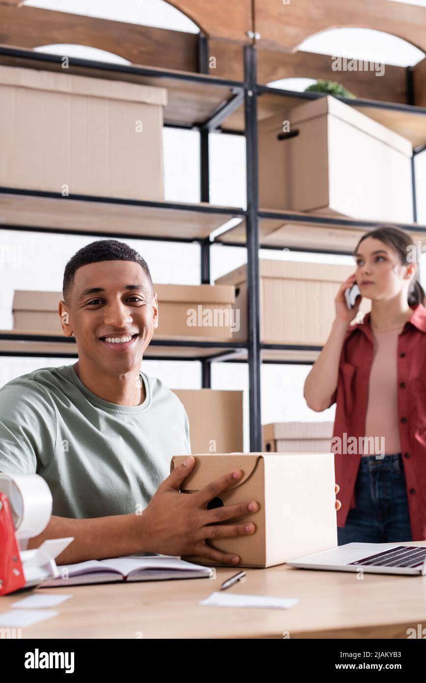 Positive african american proprietor holding carton box near laptop and ...