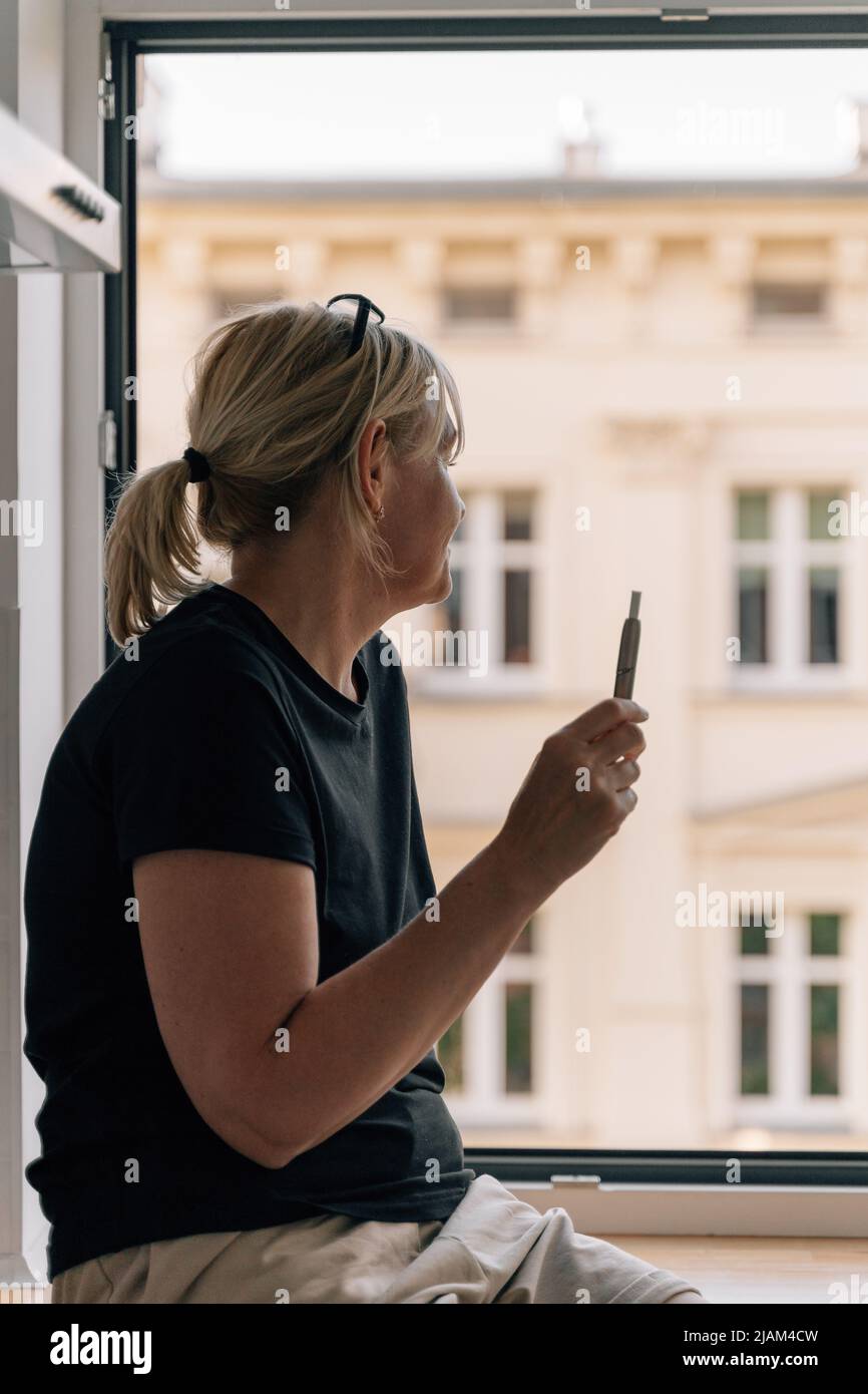 Mature woman smoking e cigarette at home near the window. Caucasian 40 ...