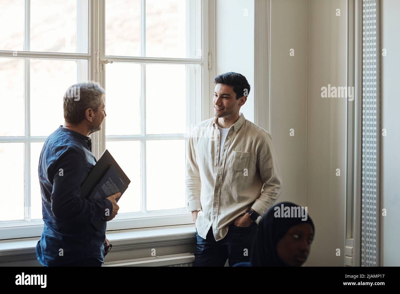Male teacher talking with student while standing by window in ...