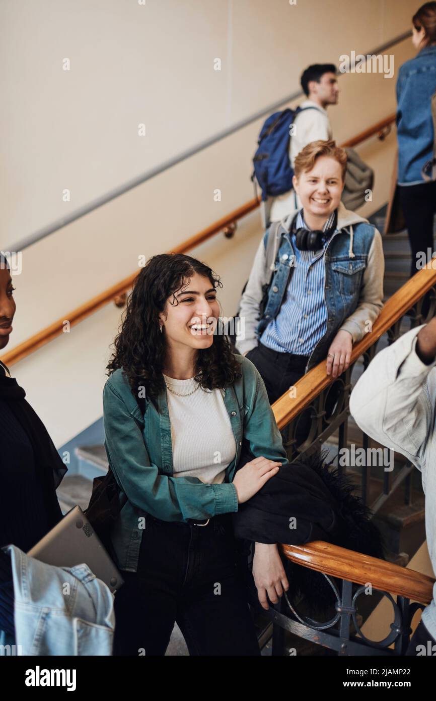 Smiling female university student with friends enjoying at staircase ...