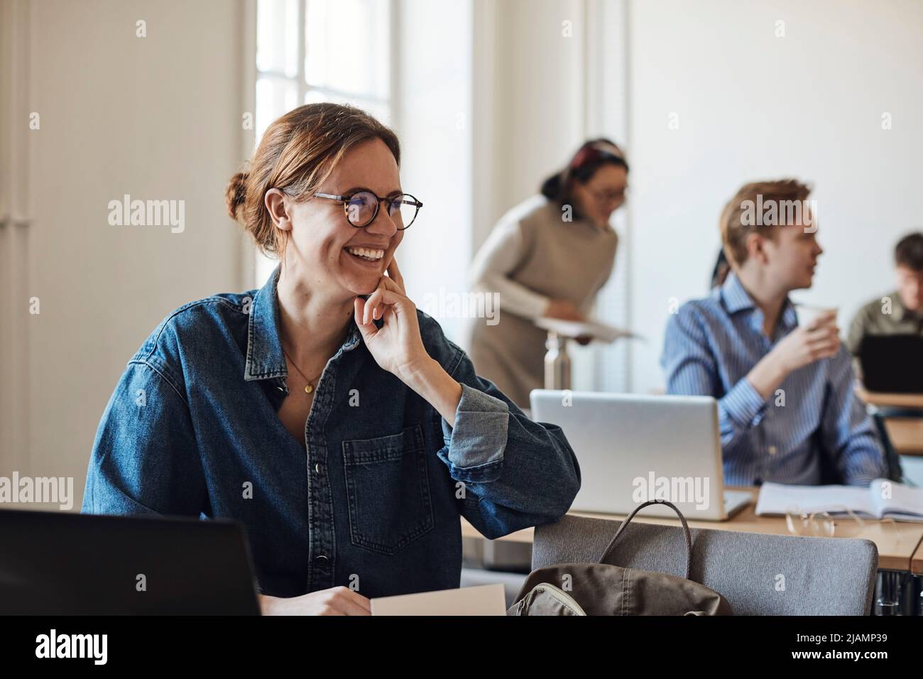 Happy female student with eyeglasses looking away while sitting in ...