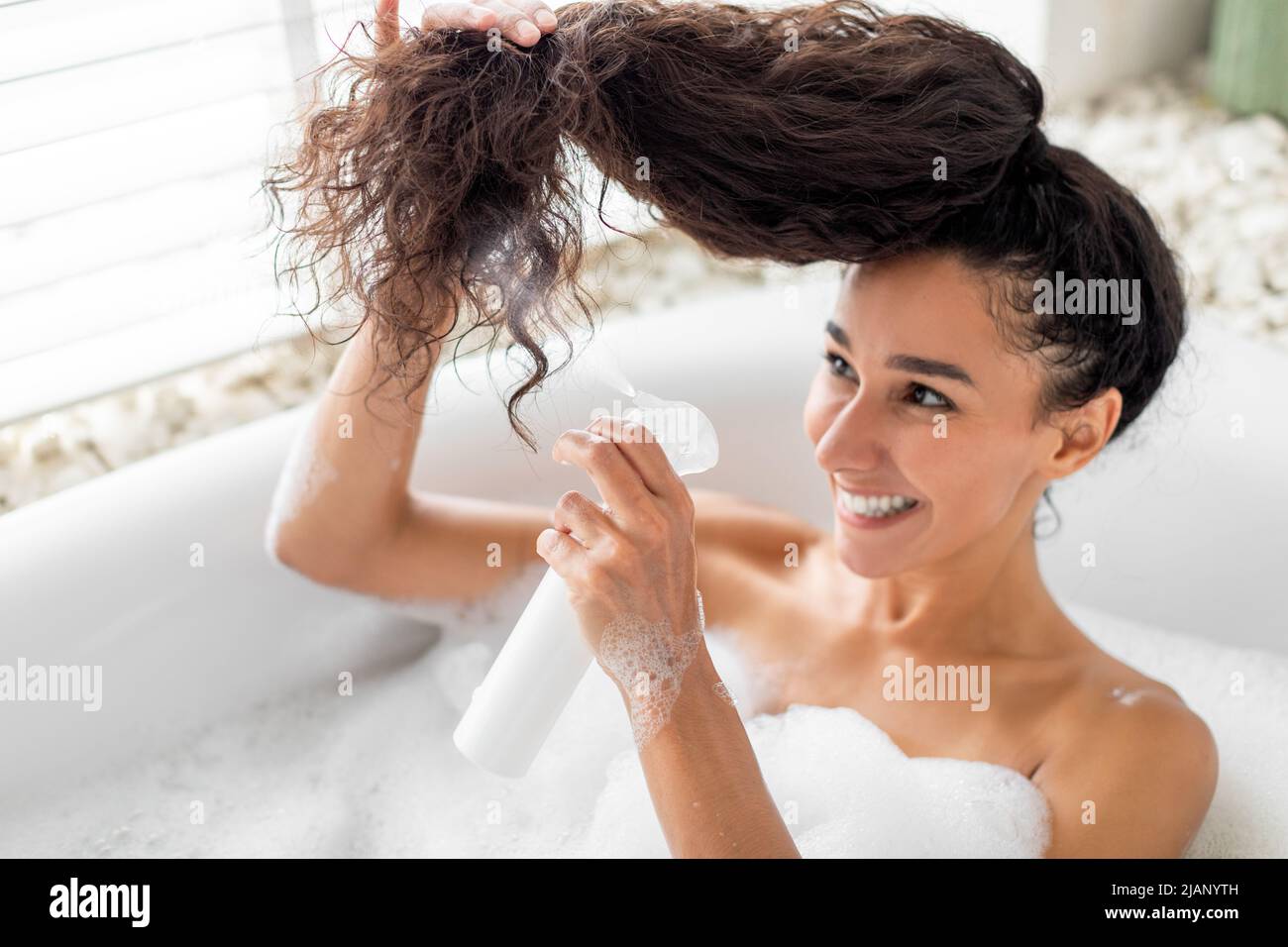 Happy young woman applying split ends repair hair spray while relaxing ...