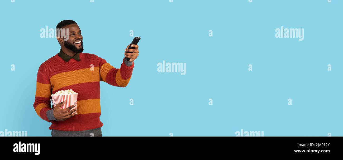 Excited young black man watching TV and eating popcorn over blue ...