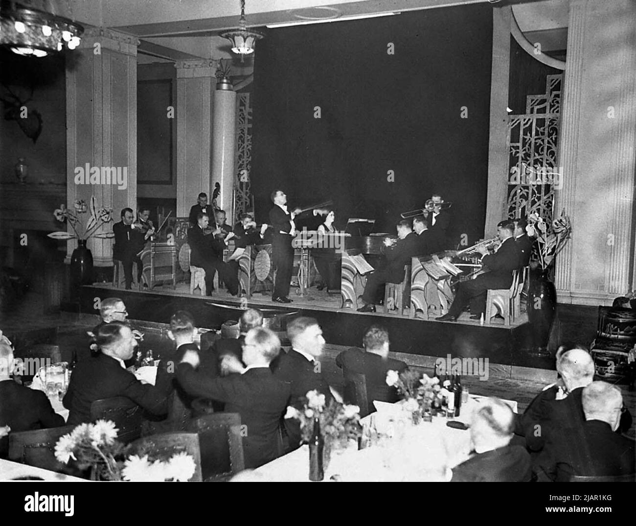 Conductor leading a band on stage in front of a group of men watching ...