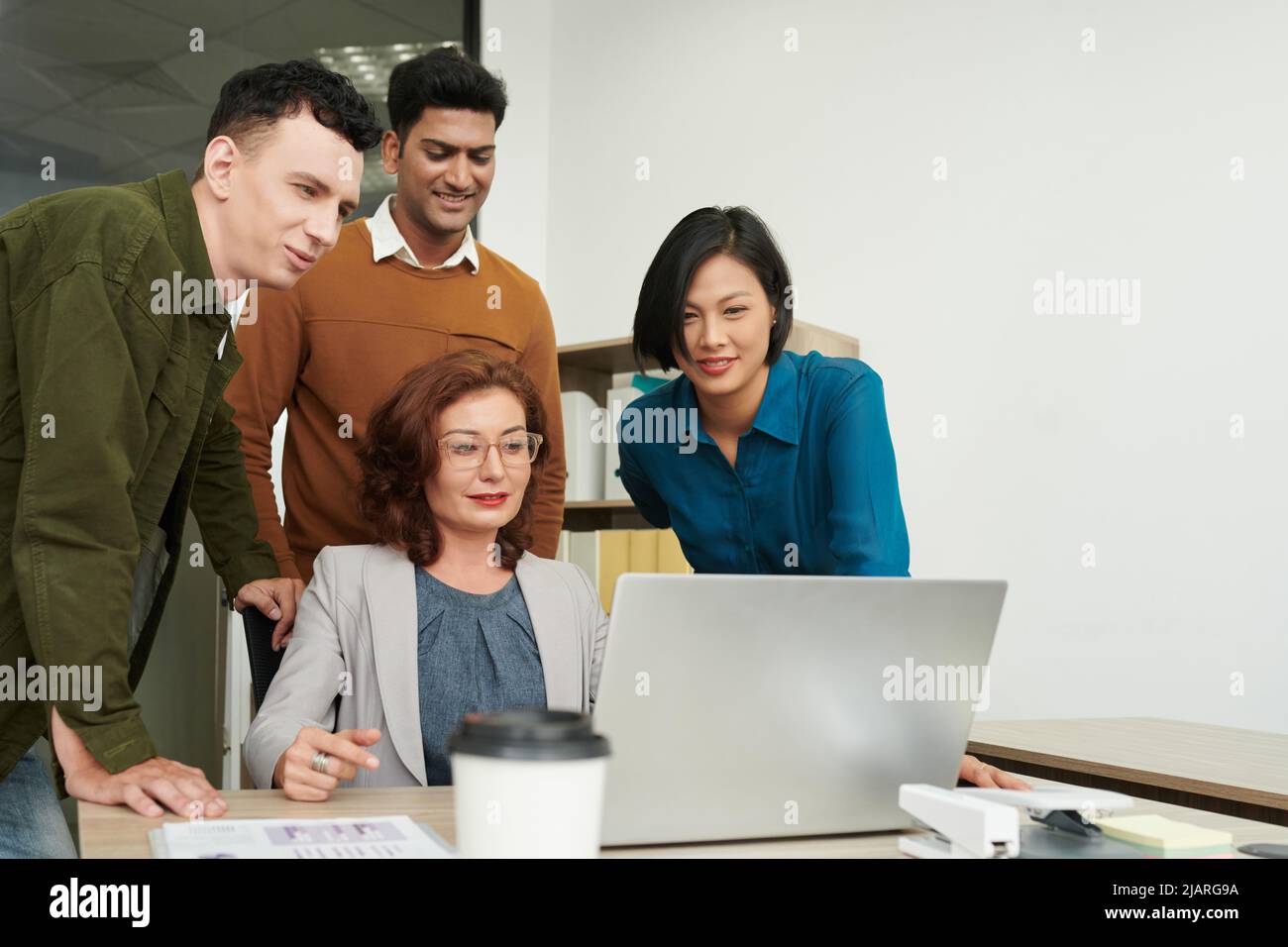 Excited coworkers gathered around desk of female entrepreneur showing ...