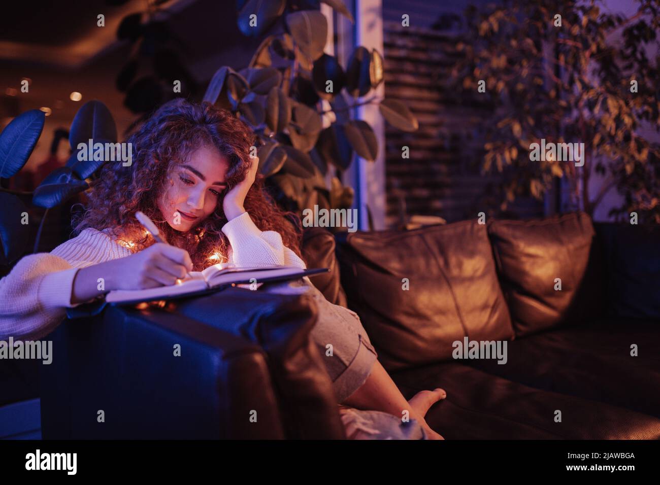 Happy young woman sitting on sofa and writing in diary in the evening ...