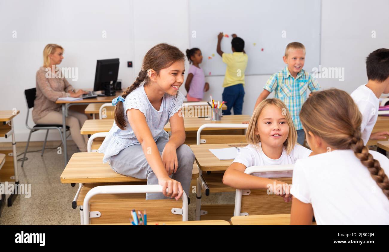 Happy preteen schoolchildren communicating during recess in classroom ...