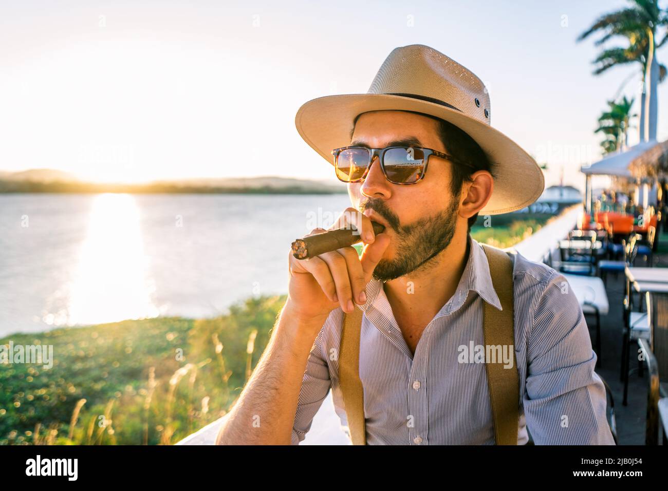 Latin hipster in a bar on the shore of a lake at sunset wearing a hat ...