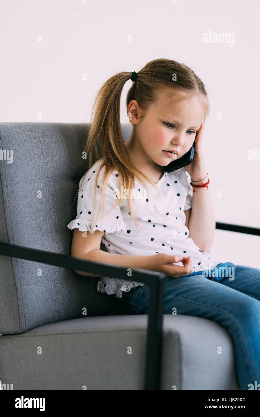 Young little confused girl talk phone sit on chair on white background ...