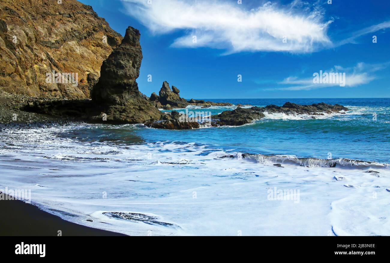 Beautiful secluded black lava sand beach, white wave foam, rock pillar, blue summer sky - Caleta ...
