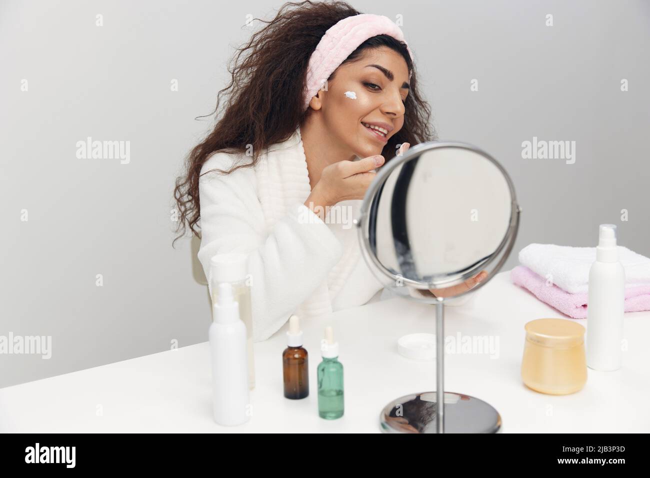 Cute tanned pretty curly Latin lady apply facial cream at cheek ...