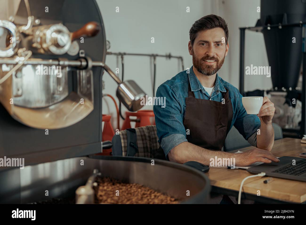 Smiling barista drinking coffee sitting near coffee roasting machine ...