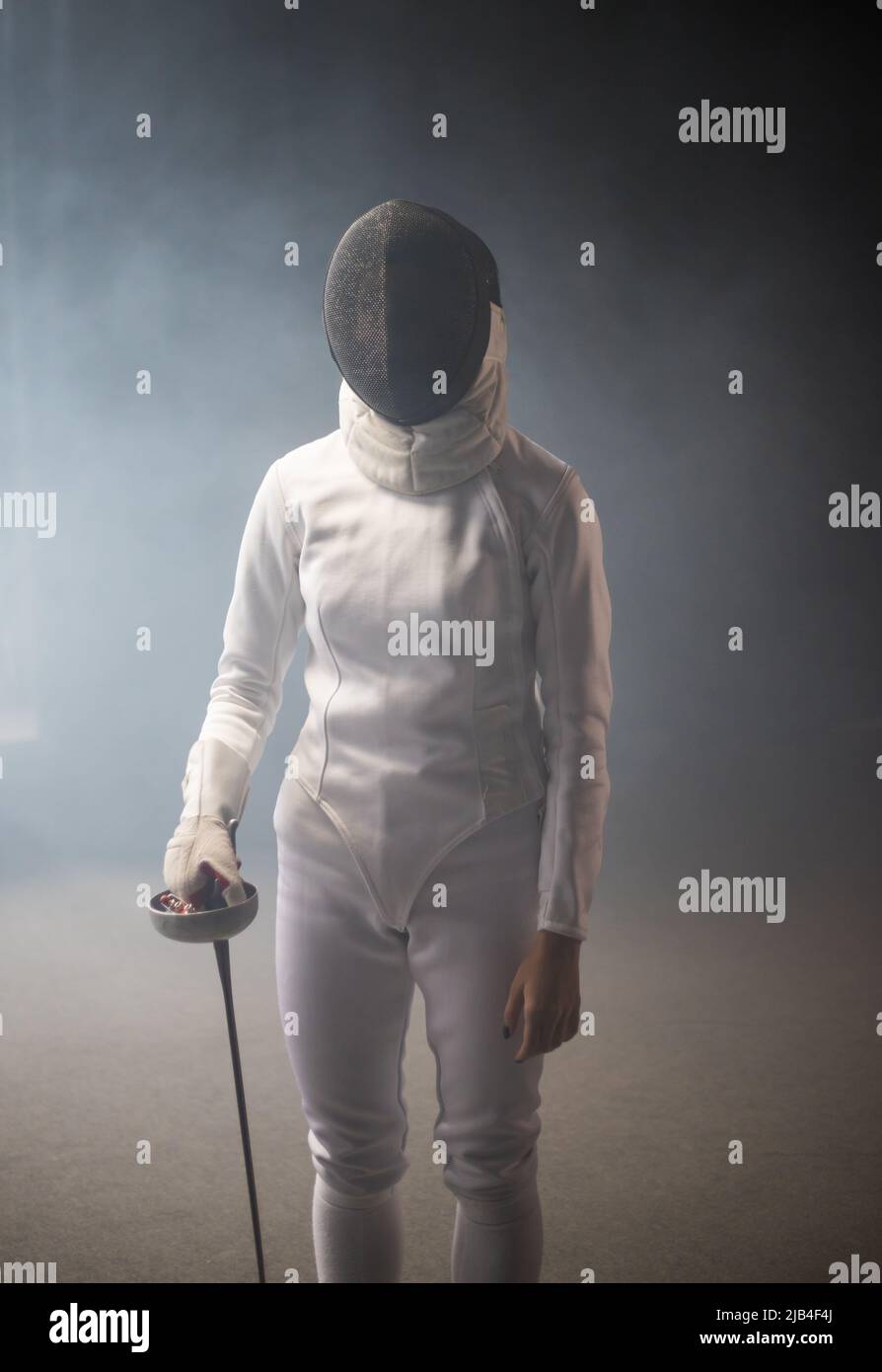 A young woman fencer in protective helmet standing with a sword down ...