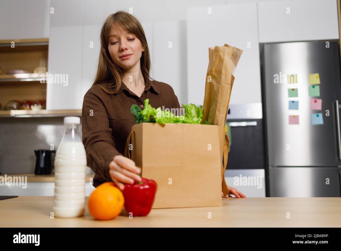 Young woman unpacking vegetables and other food from paper grocery bag ...
