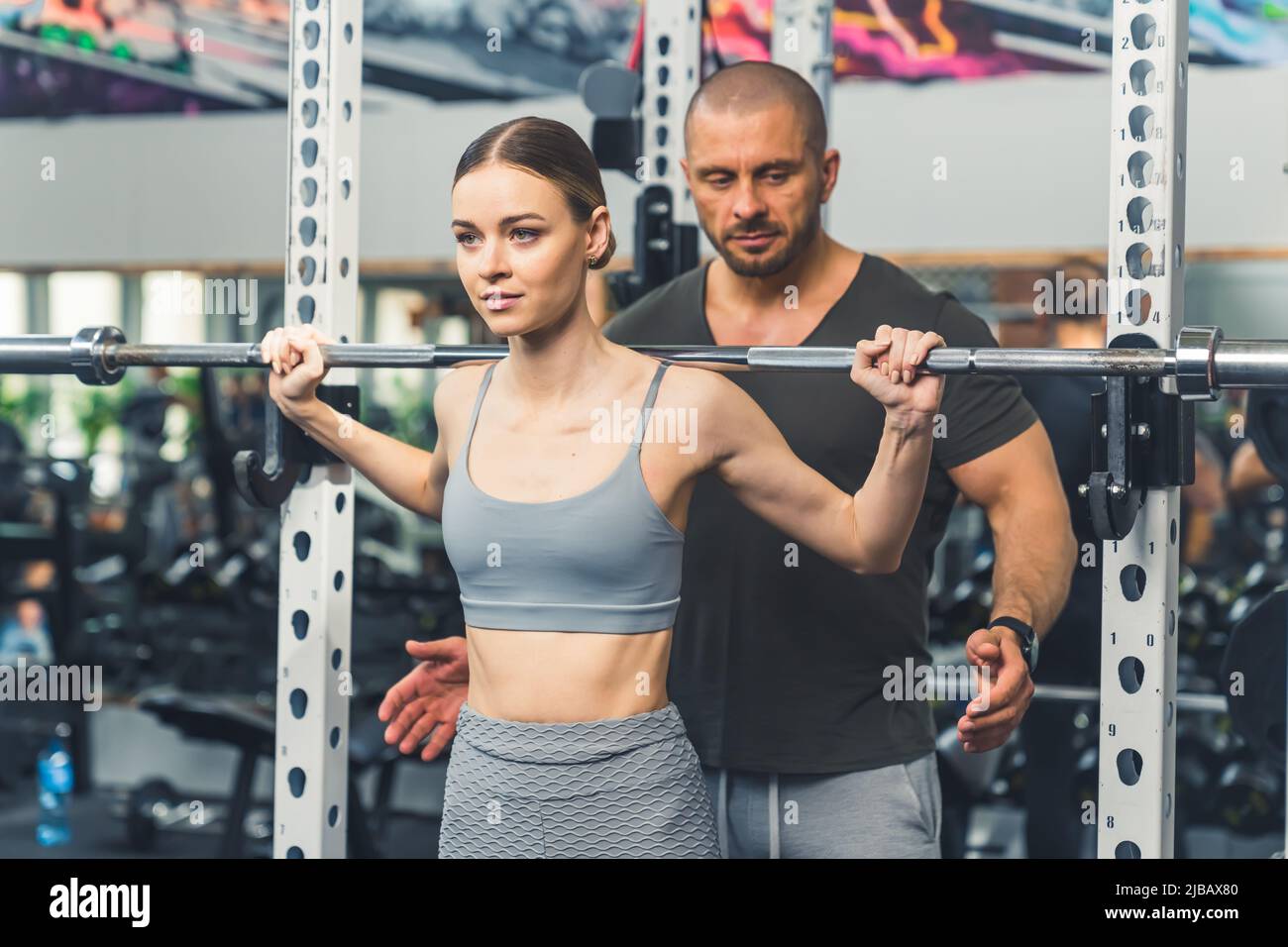 Shoulder barbell lifting. Confident caucasian girl trying out weightlifting under the guidance ...