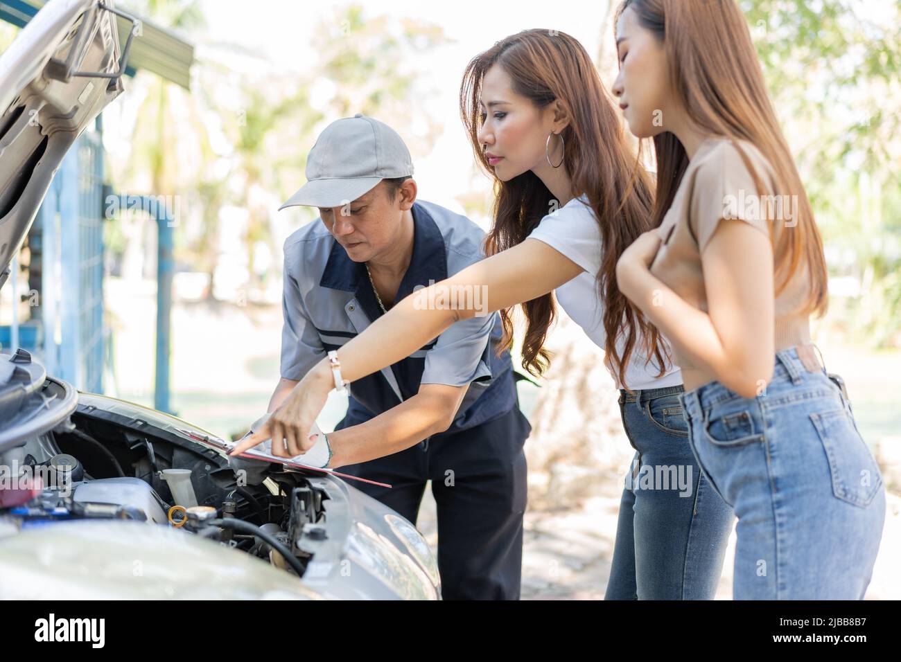 Asian male auto mechanic holds a clipboard and examines car engine ...