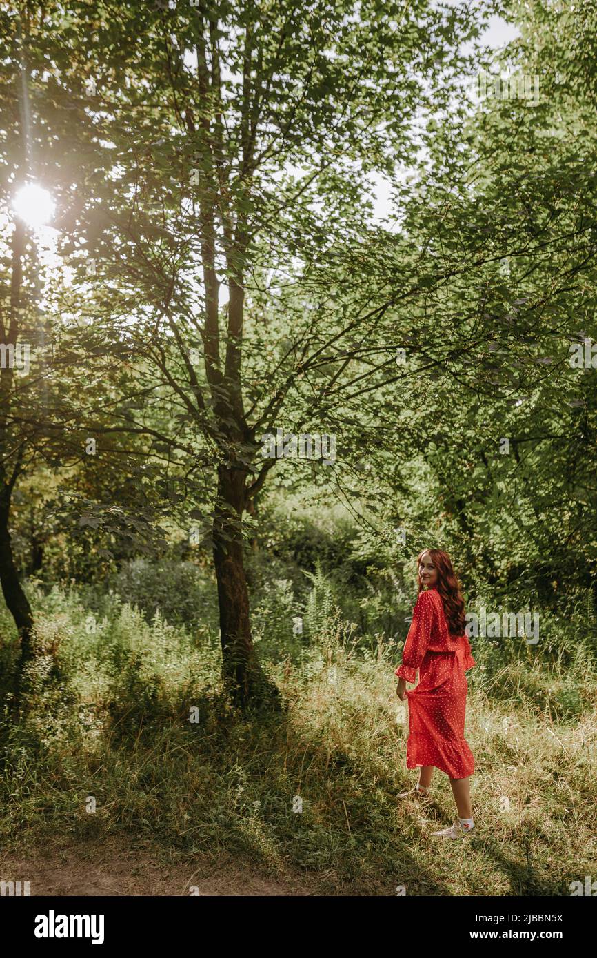 Pretty young redhead woman in red dress walking through meadow with ...