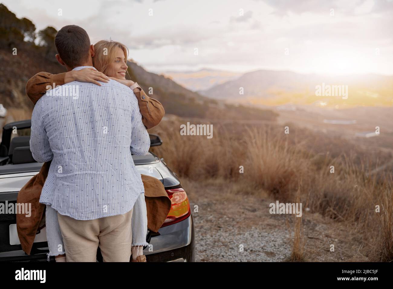 Rear of a man hugging his girlfriend outdoors at sunset, mountains view ...