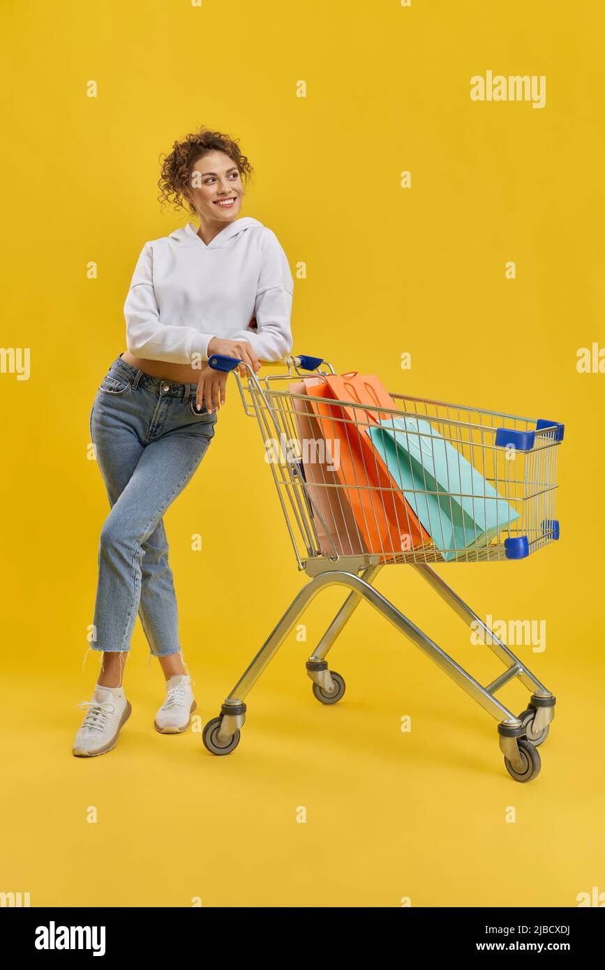 Cute girl in casual clothes, leaning on cart with purchases in studio ...