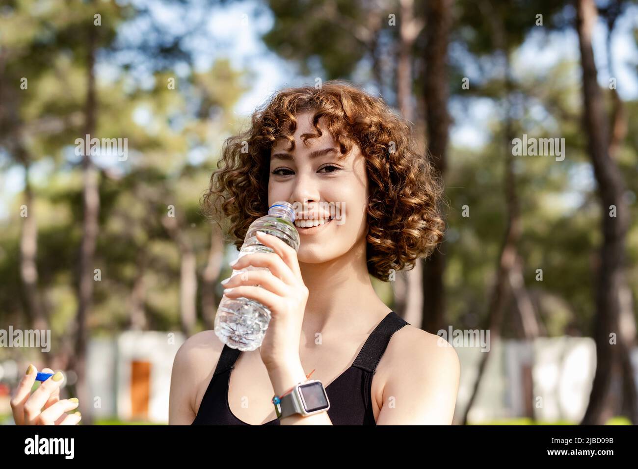 Cheerful redhead woman wearing sports bra standing on city park ...