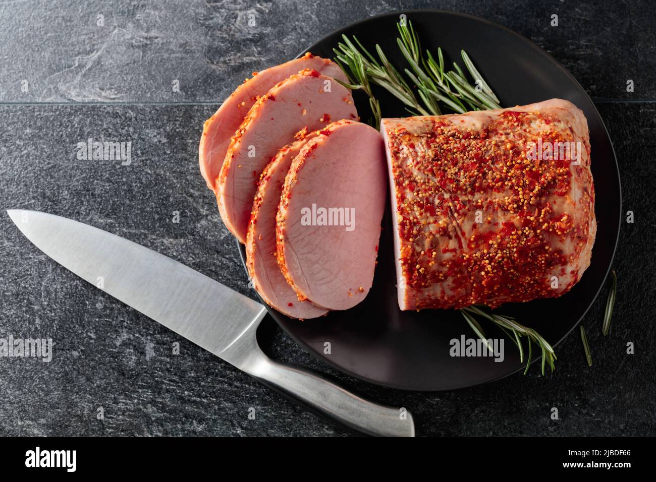 Spicy smoked ham with rosemary and kitchen knife on a black stone table ...
