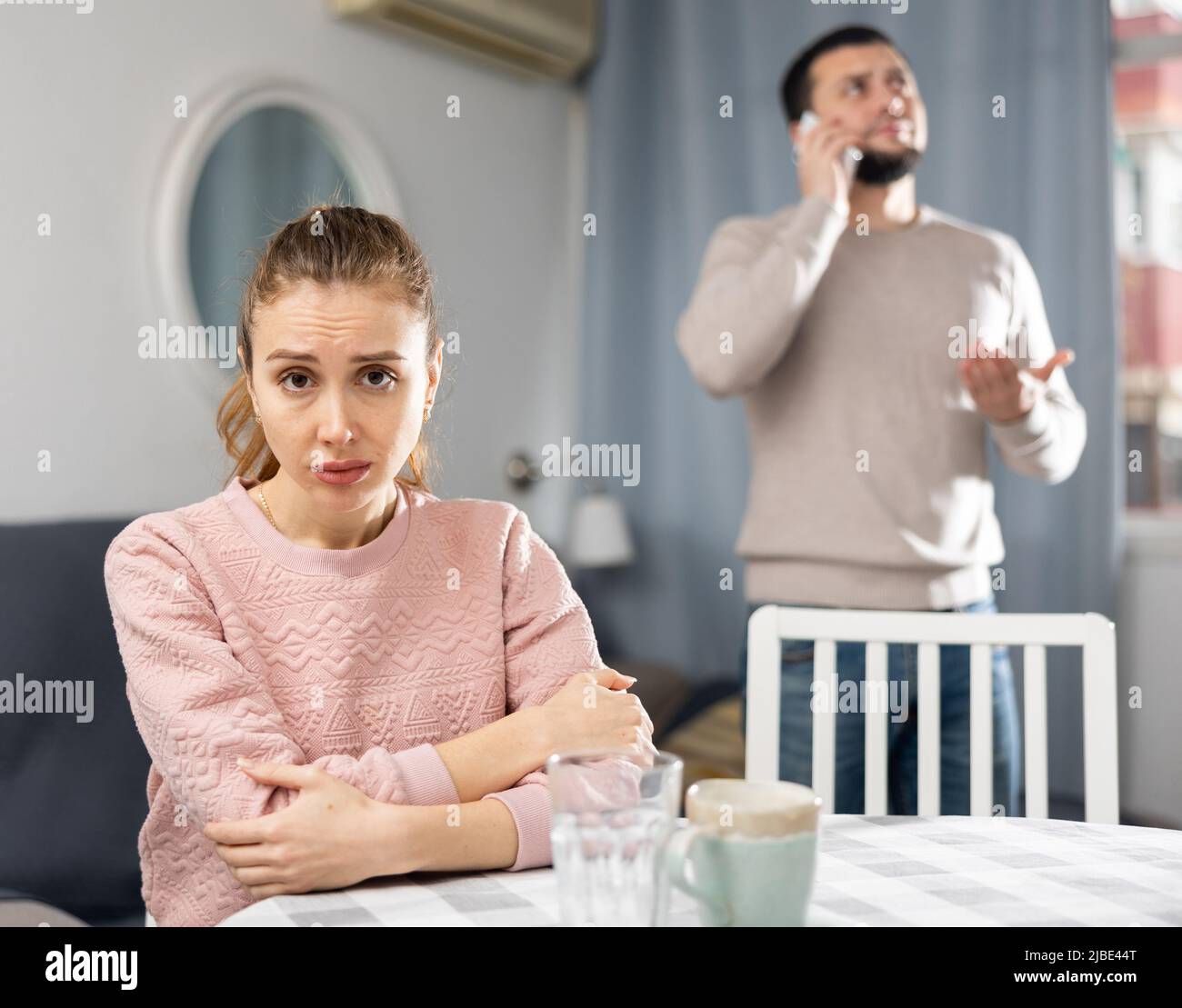 Sad woman sitting at table at home, husband talking on phone Stock ...