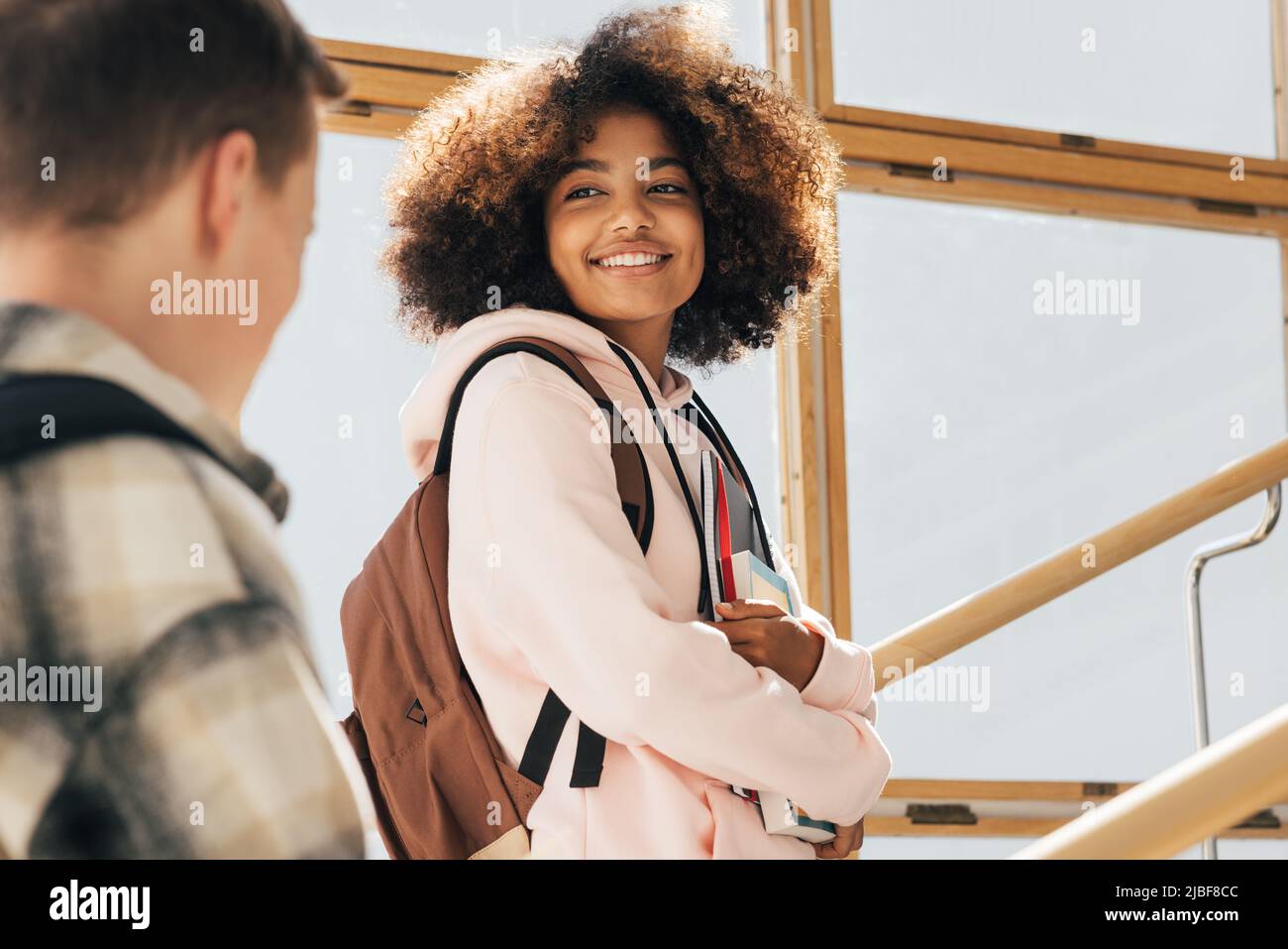 Smiling girl with books and backpack looking at classmate while walking ...