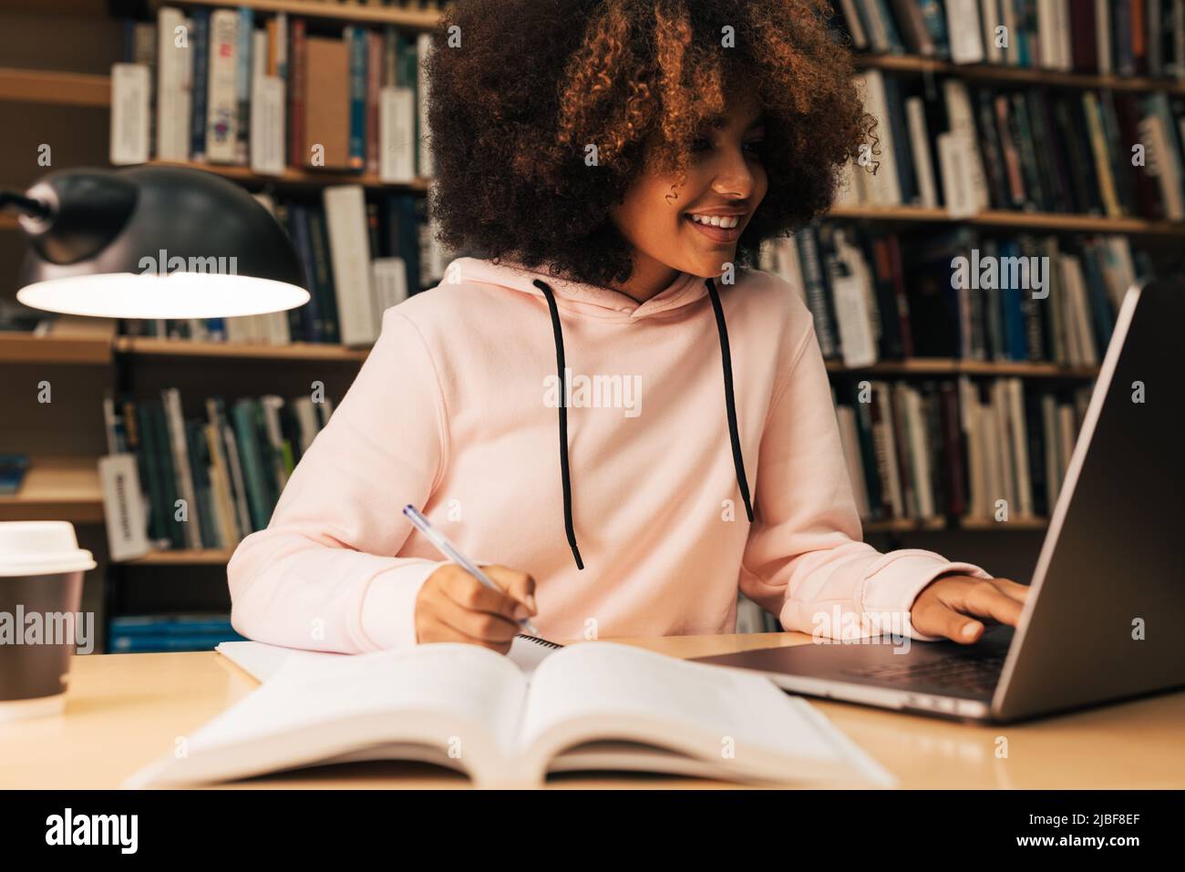 Smiling girl taking notes in her text book under lamp in library. Young ...