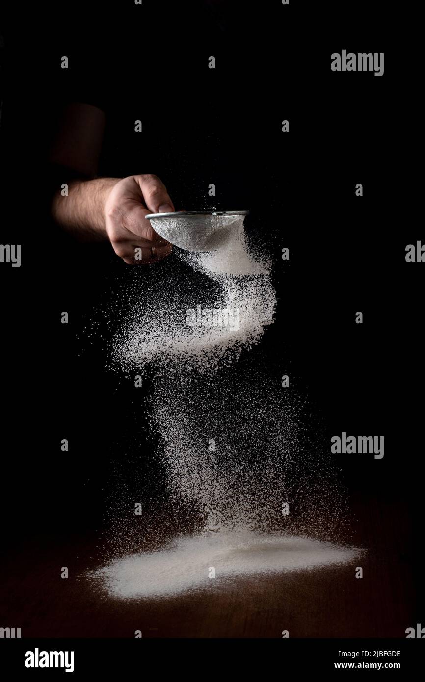 Flour flies through a sieve on a black background. A man sifts flour ...