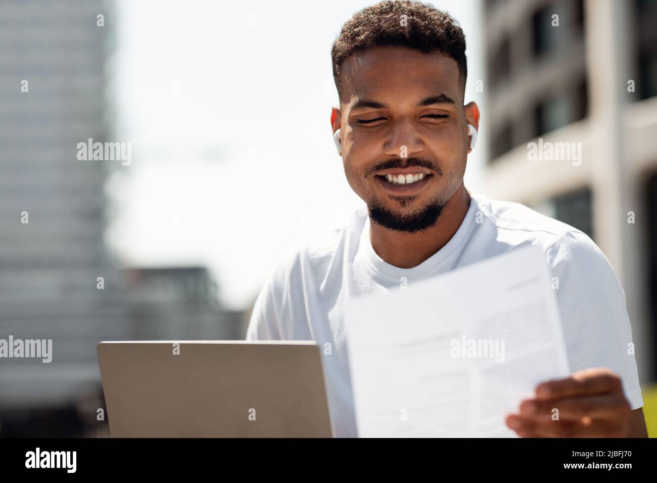 Happy black man working with documents and laptop computer outdoors ...