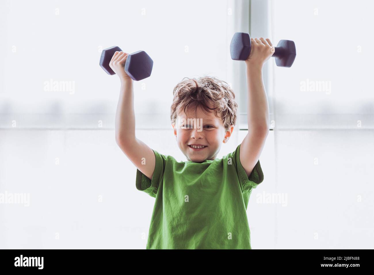 Happy kid in activewear smiling and exercising with dumbbells against ...