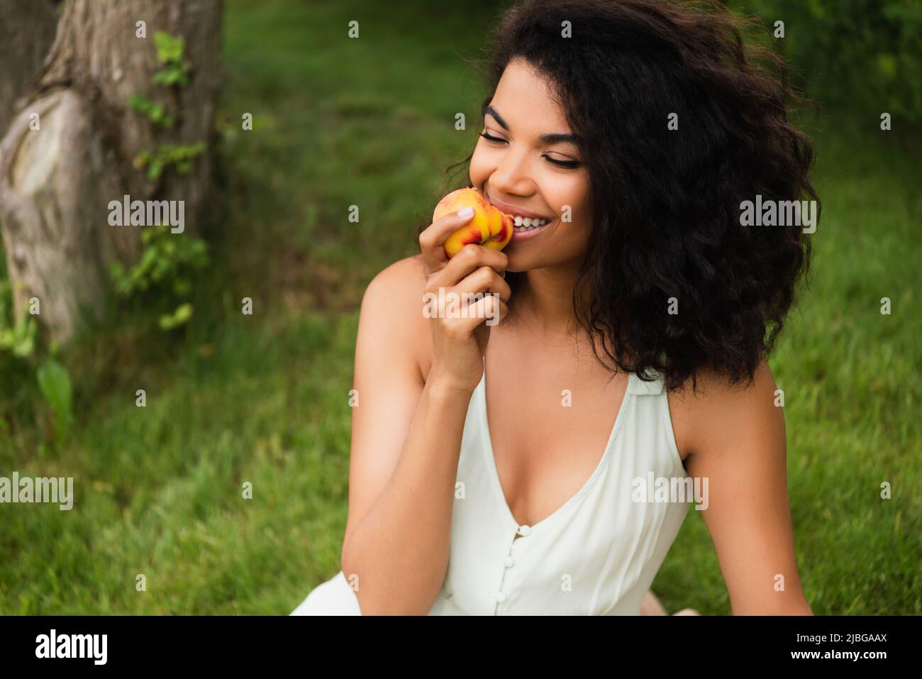 happy and young african american woman in white dress eating peach in ...