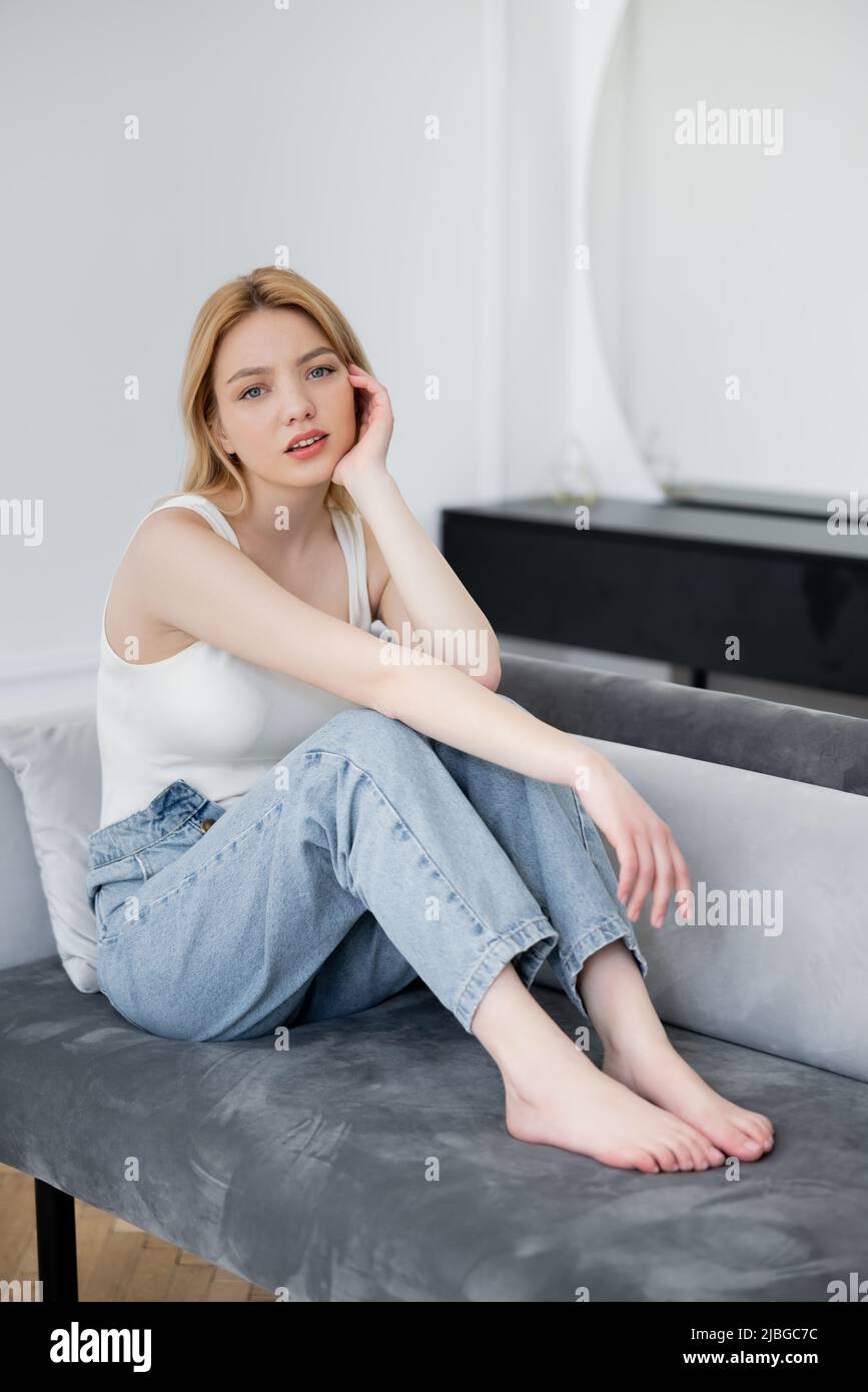 Barefoot woman in jeans looking at camera while sitting on couch at ...