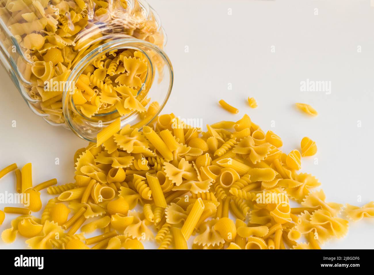 Uncooked, dry assorted pasta pouring out of glass jar, on white surface ...