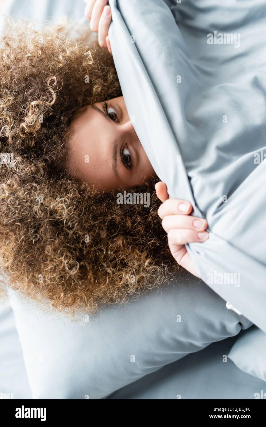 top view of curly woman obscuring face with blanket and looking at ...