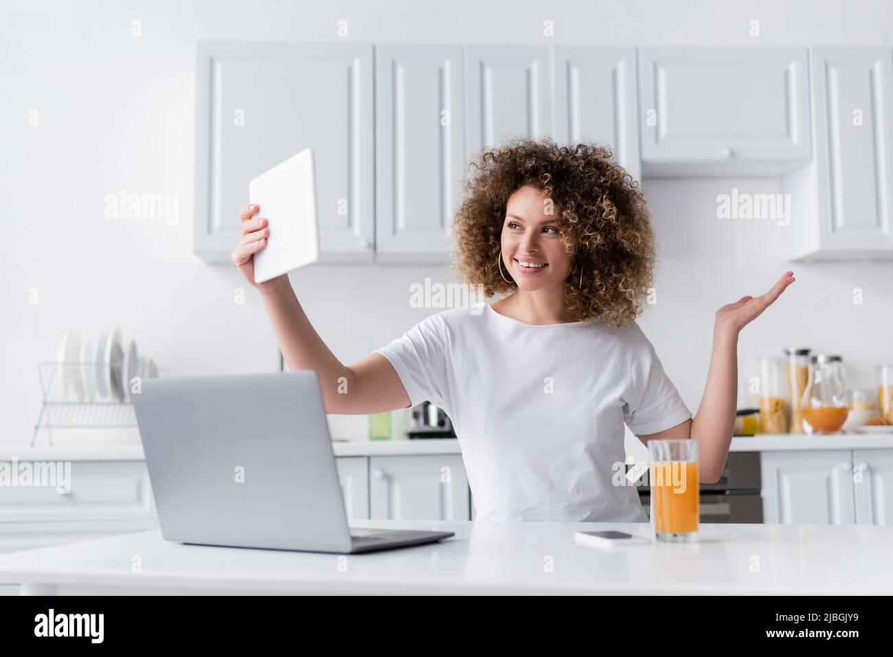 cheerful woman with wavy hair taking selfie on digital tablet near ...
