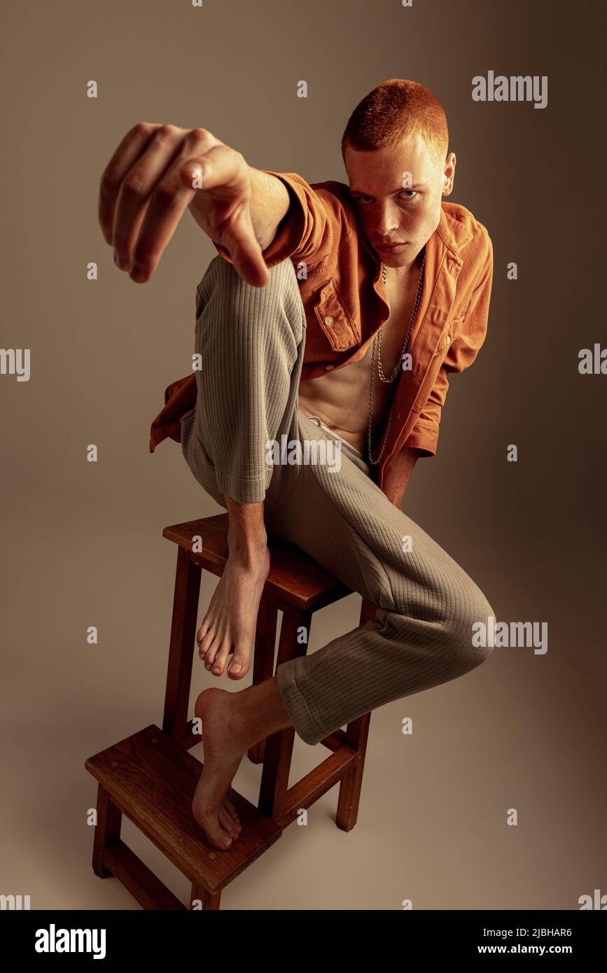 Stylish red-haired young man sitting, posing isolated over brown studio ...