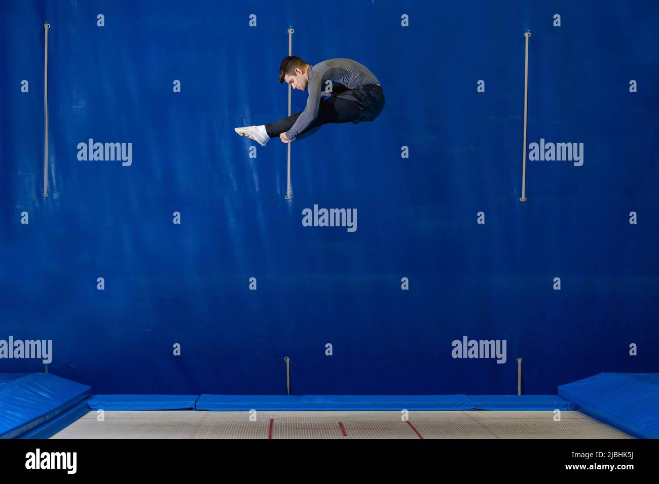 Young man doing an acrobatic figure on elastic bed, in blue background ...