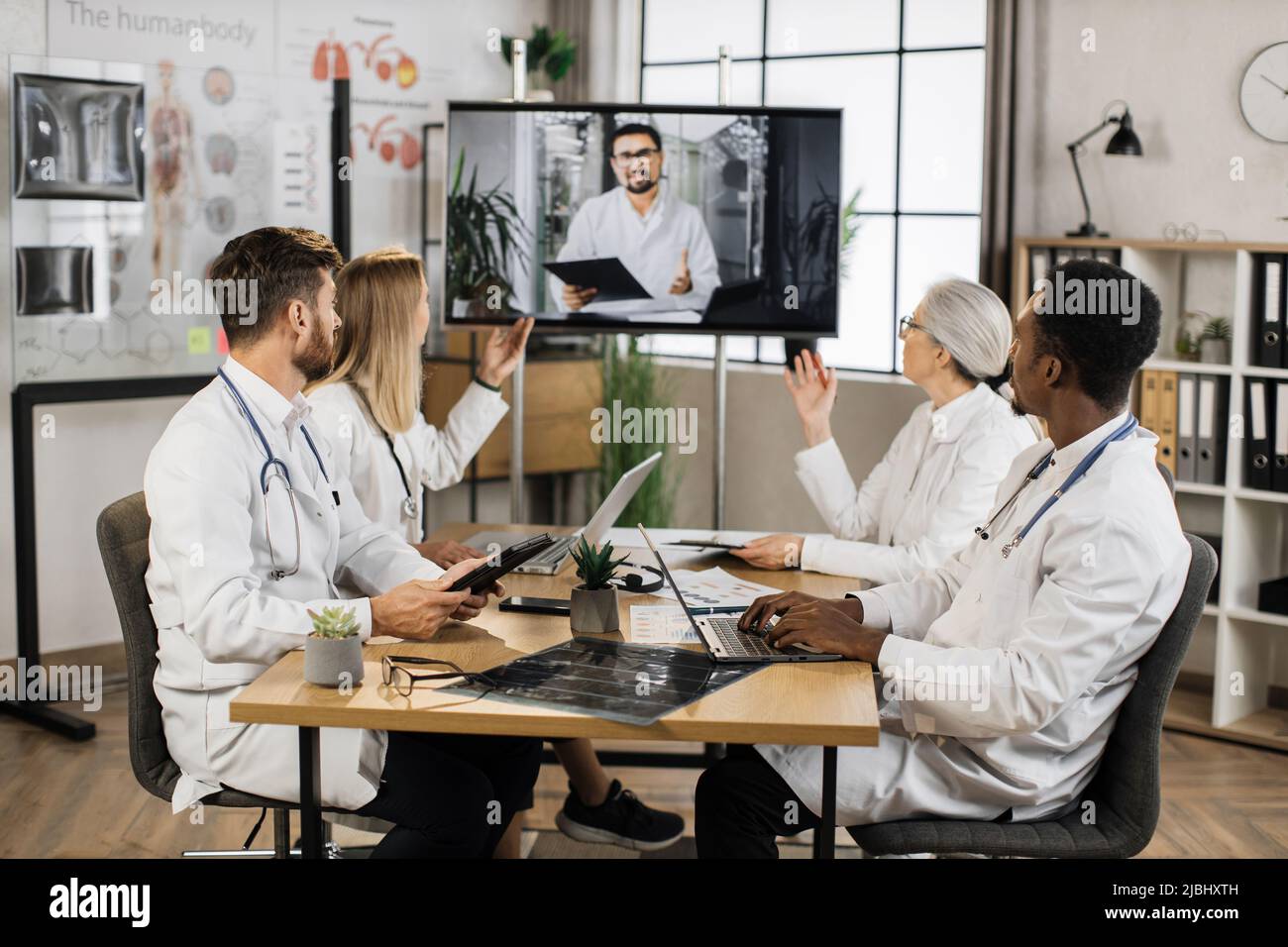 Four multiracial doctors of various ages sitting at desk at boardroom ...