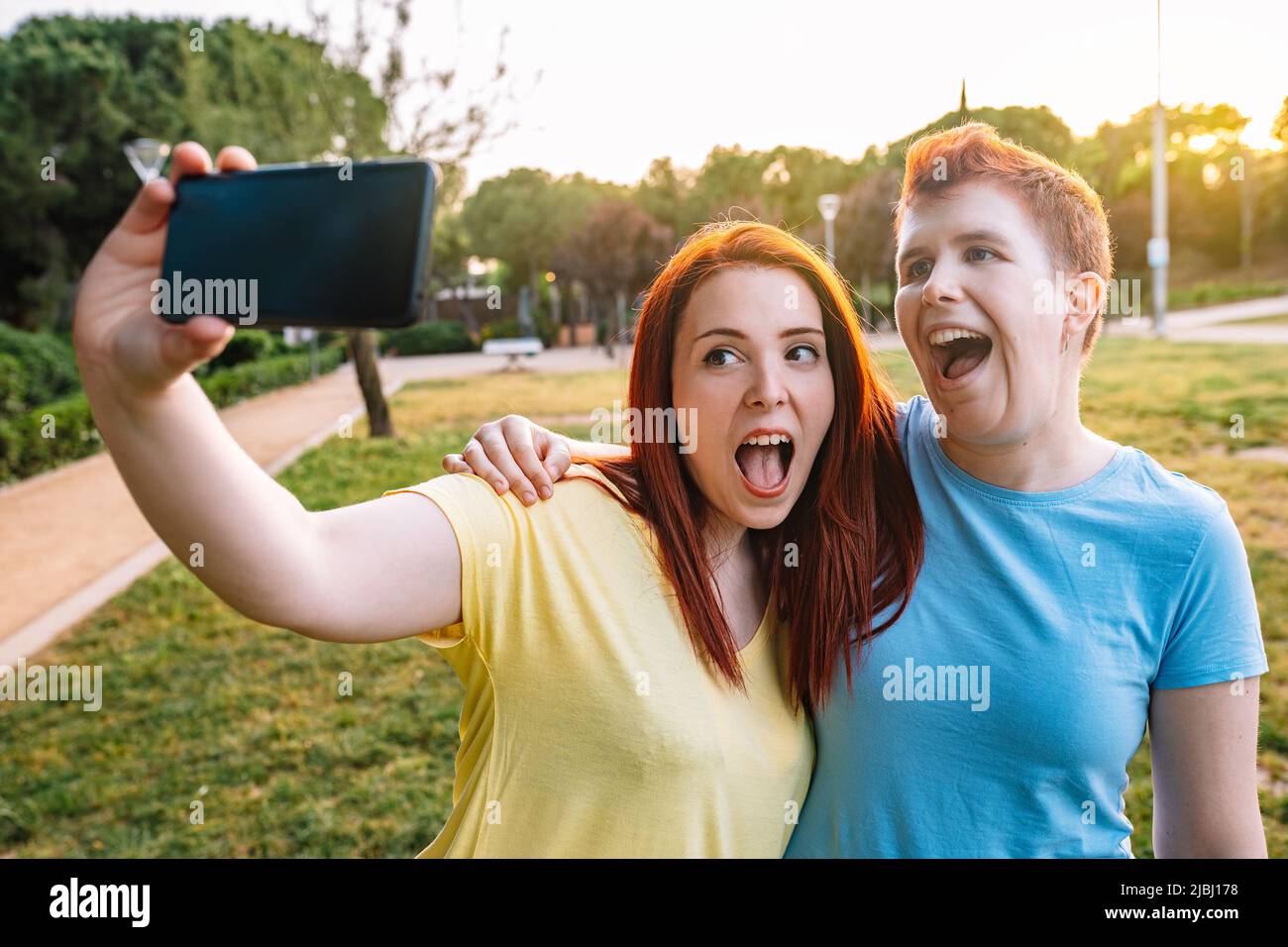 two friends taking a selfie with their smartphones, making funny faces in a public city park at ...