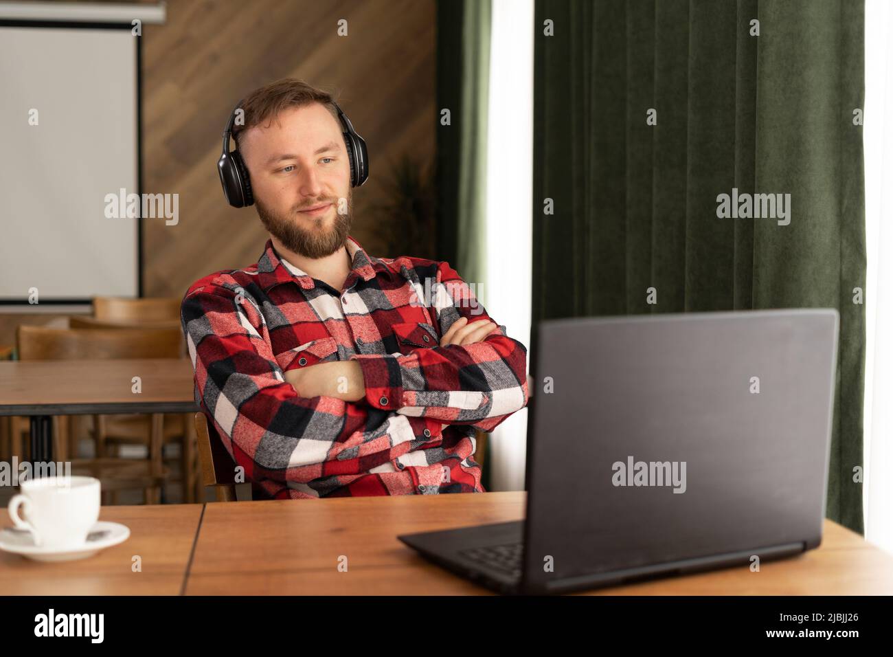 Pensive bearded man in headphones sitting on publicity area with laptop ...