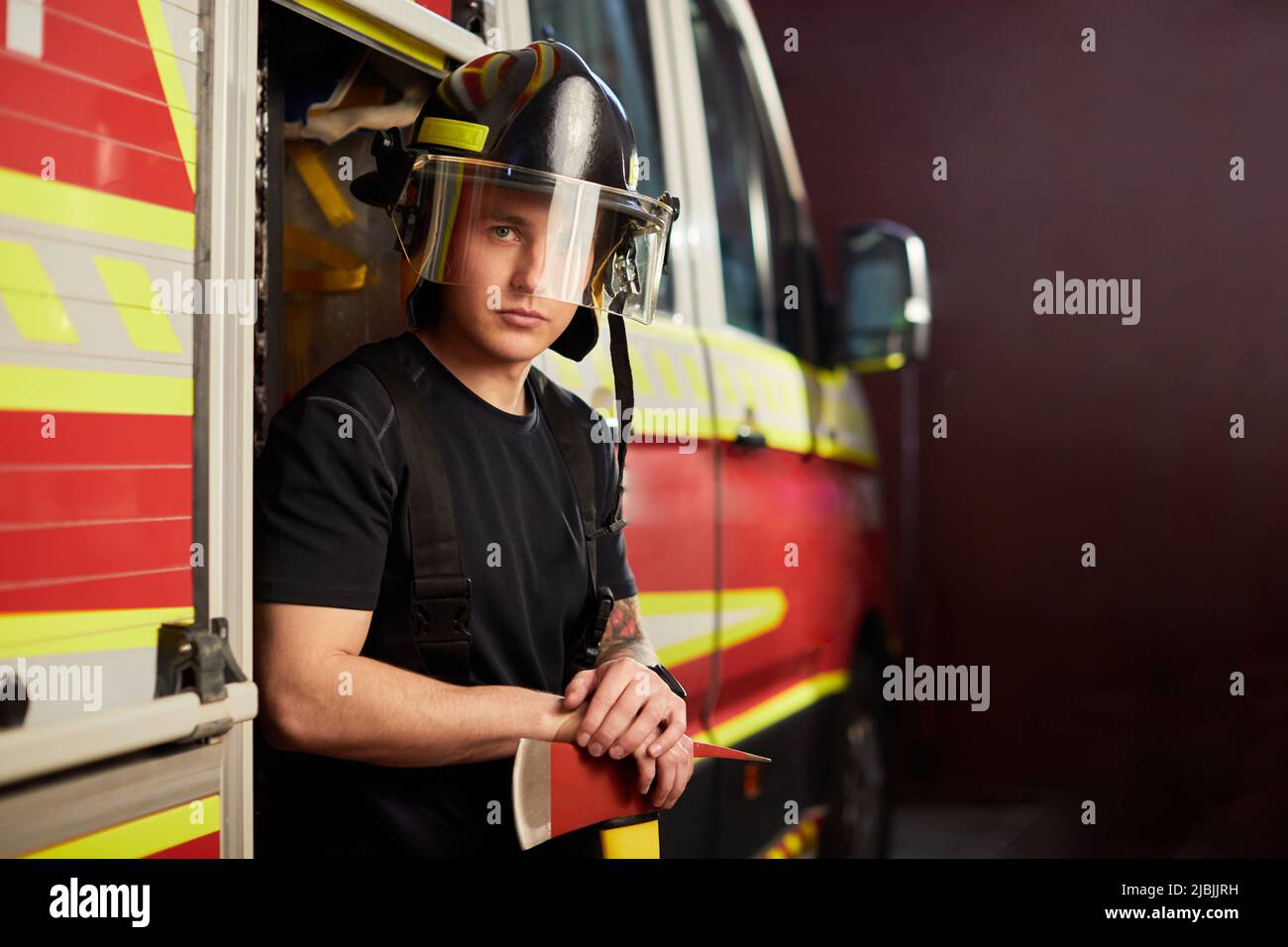Photo of fireman wearing helmet with ax against fire engine Stock Photo ...