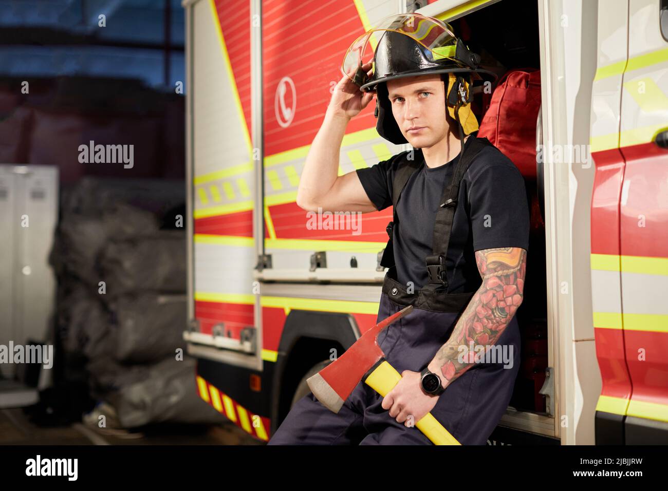 Photo of fireman wearing helmet with ax against fire engine Stock Photo ...